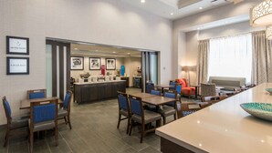 Um saguão de hotel com balcão de recepção, área de refeições com mesas e cadeiras, e um espaço de estar com sofá e poltronas.