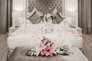 Quarto de hotel com uma cama grande, duas mesas de cabeceira, um painel de cabeceira estofado e um arranjo floral sobre a cama.