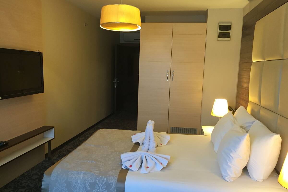 Quarto de hotel com cama, televisão de tela plana na parede, mesa de cabeceira, abajur e guarda-roupa.