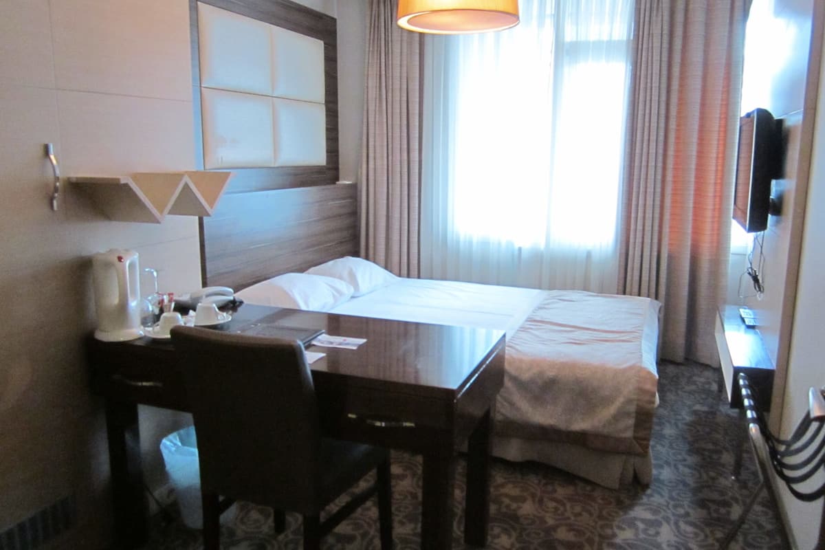 Quarto de hotel com cama, escrivaninha, cadeira, luminária, televisão e janela com cortinas.