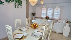 Uma sala de jantar com uma mesa branca posta para quatro pessoas, rodeada por cadeiras brancas. Um lustre pendurado acima e duas janelas com persianas brancas.