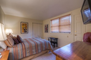 Quarto com cama, escrivaninha, cadeira e televisão fixada na parede.