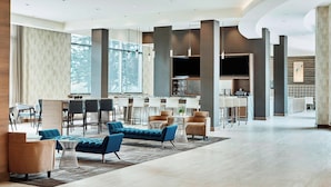 Um hall amplo com design moderno, com área de estar com sofás e cadeiras, área de refeições com mesas e cozinha ao fundo.