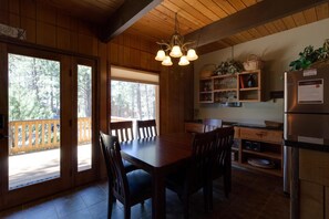 Uma cabana de madeira com mesa e cadeiras, uma cozinha com prateleiras e vista para a floresta através de grandes janelas.