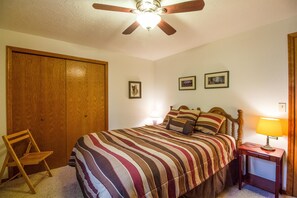 Quarto com cama, guarda-roupa de madeira, cadeira, mesa de cabeceira com abajur e ventoinha no teto.