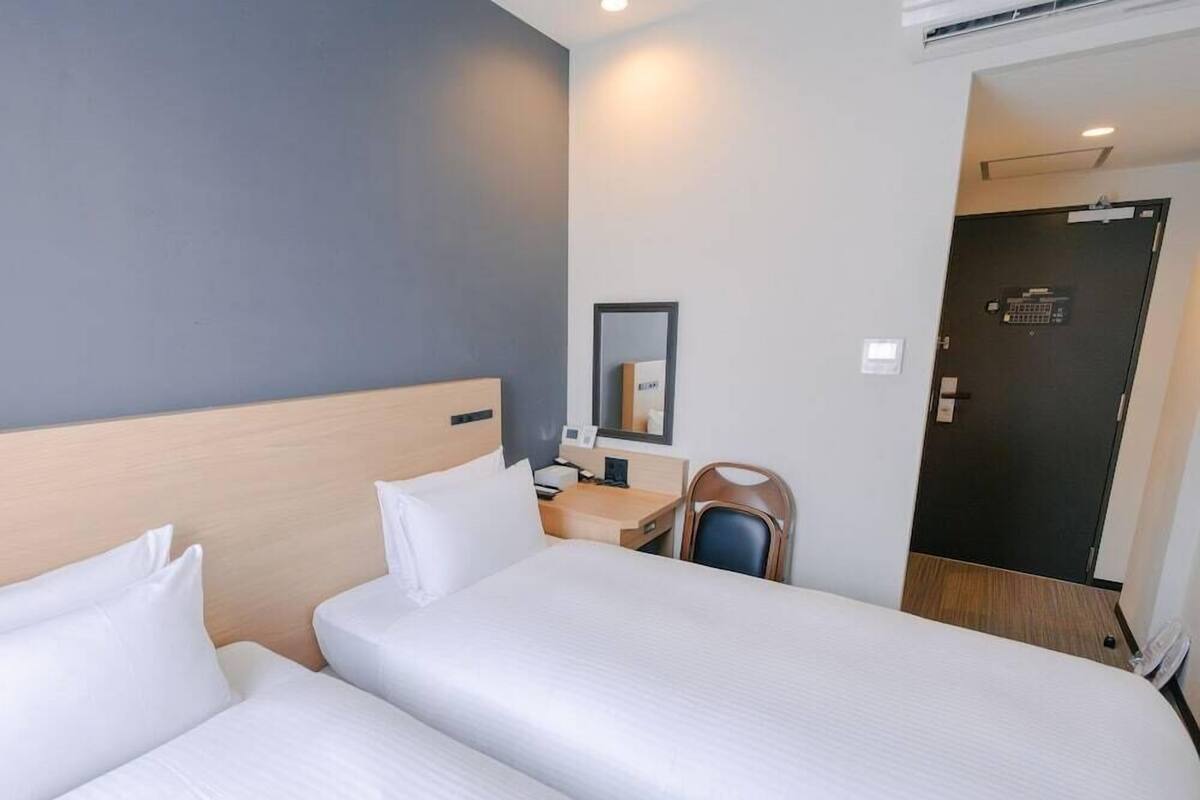 Quarto de hotel com duas camas, uma mesa, uma cadeira, um espelho e uma porta.