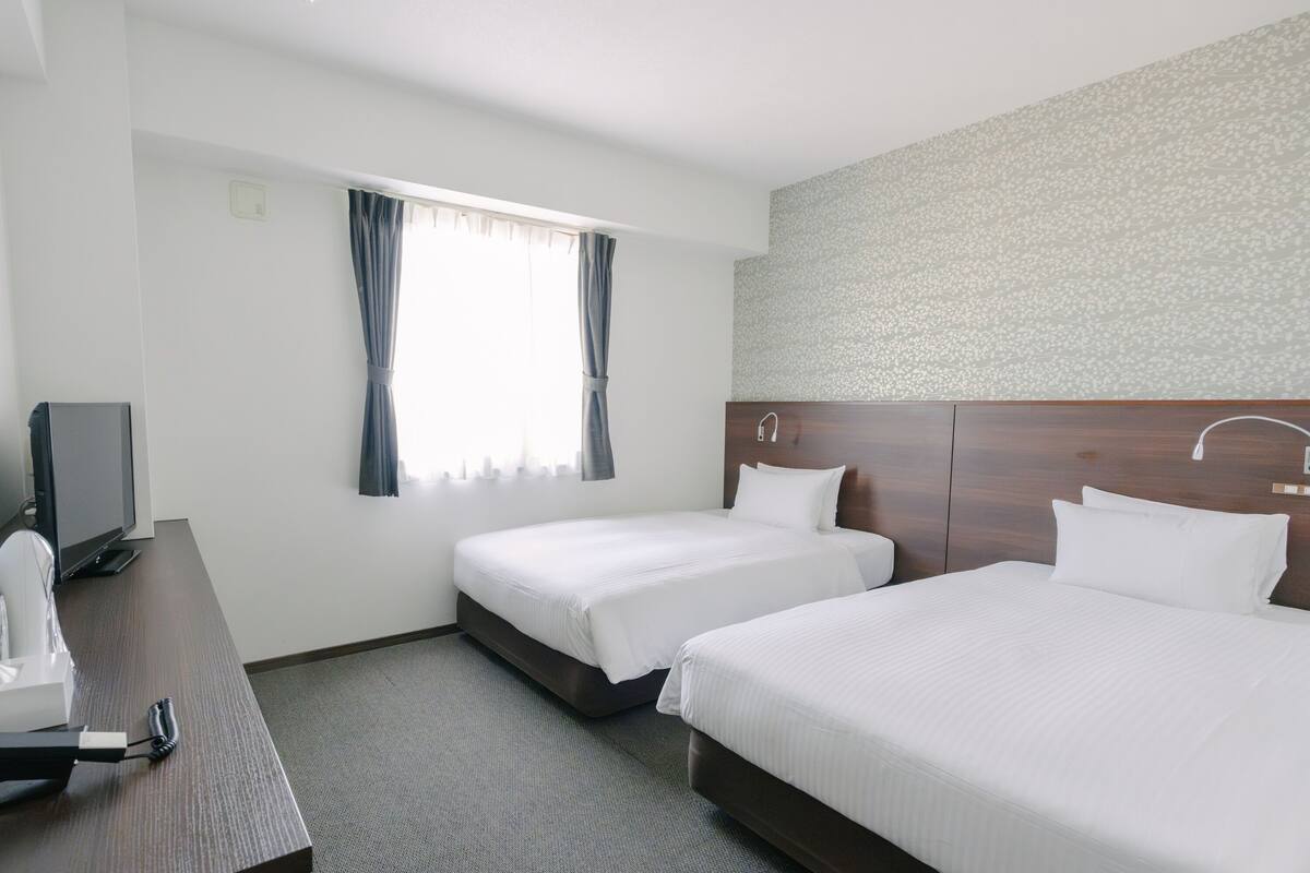Quarto de hotel com duas camas, uma mesa de trabalho com televisão e uma janela com cortinas.