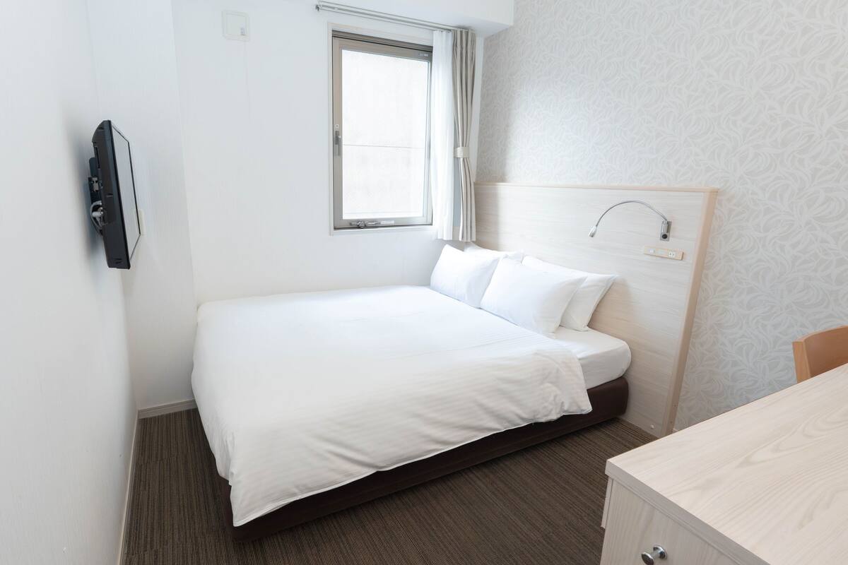Quarto de hotel com uma cama de solteiro, televisão na parede, janela com cortinas e cabeceira de madeira.