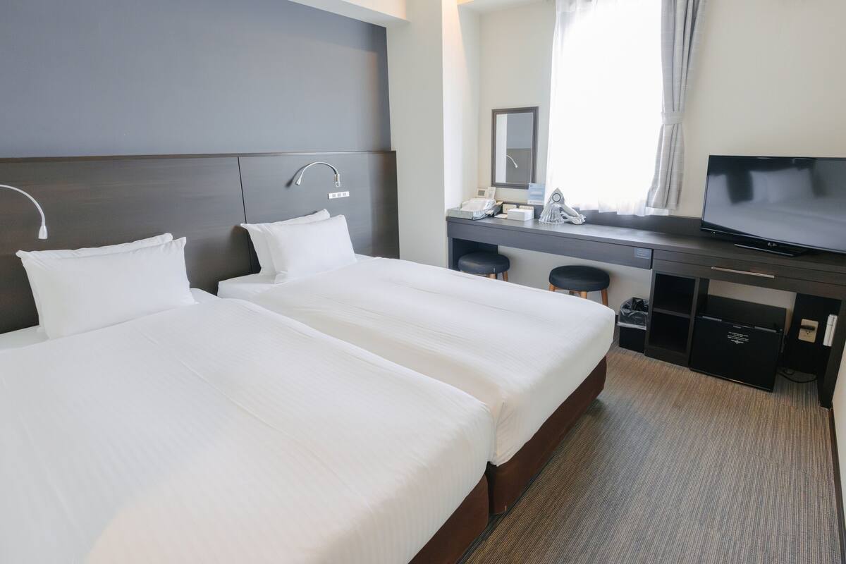 Quarto de hotel com duas camas, uma mesa de trabalho com televisão, espelho e janela com cortinas.