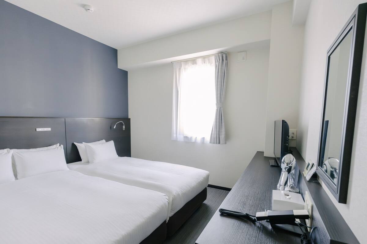 Quarto de hotel com duas camas, uma mesa de trabalho com telefone e uma janela com cortinas.