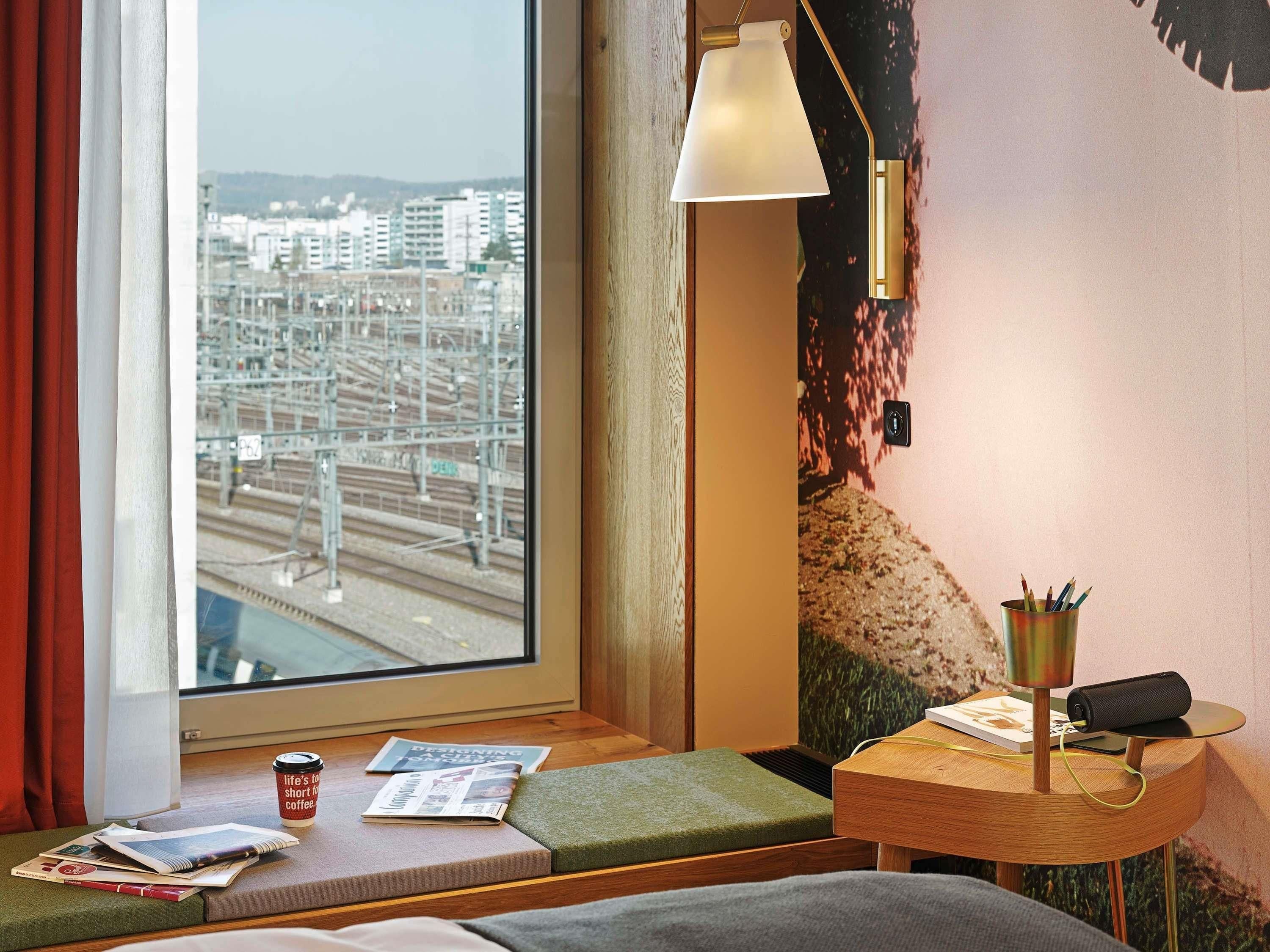 Quarto de hotel com uma janela grande, uma escrivaninha com luminária, uma xícara de café e uma revista.