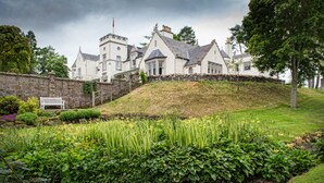 Um edifício histórico com parede de pedra, um pequeno lago e vegetação exuberante.