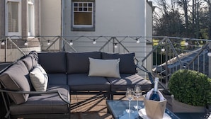 Um terraço no telhado com um sofá preto para área externa, uma mesa de vidro com copos de vinho e uma garrafa, e vista para um prédio branco com chaminé.