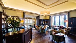 Uma área de bar com balcão de madeira, banquetas e um vaso grande com flores. Há várias áreas de estar com cadeiras e sofás, e uma janela grande com cortinas.
