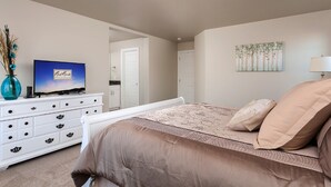 Um quarto com uma cama, cômoda, televisão e porta que dá para outro espaço.
