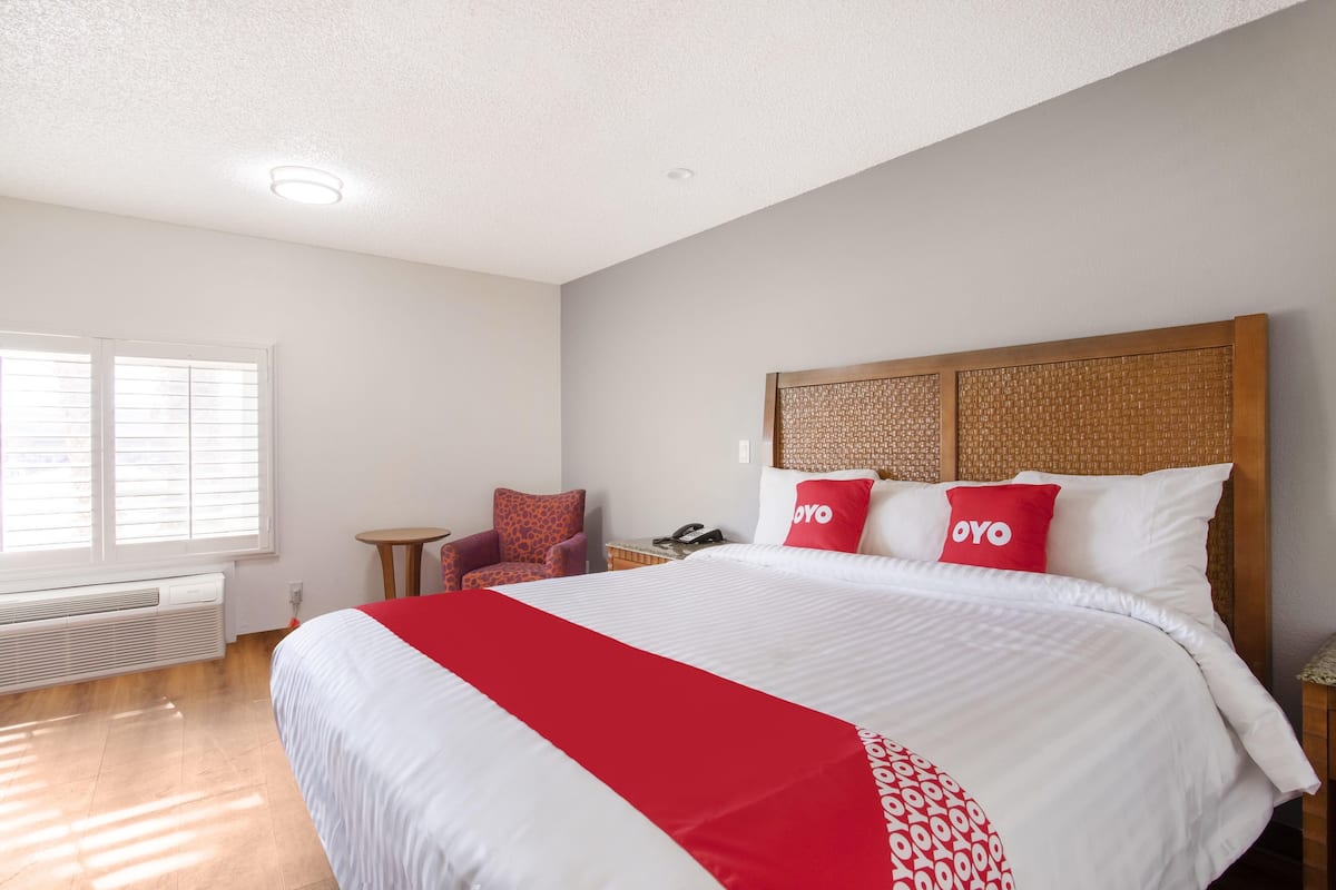 Quarto de hotel com uma cama grande, colcha vermelha e branca, cabeceira de madeira, mesa redonda pequena, uma cadeira e uma janela com persianas.