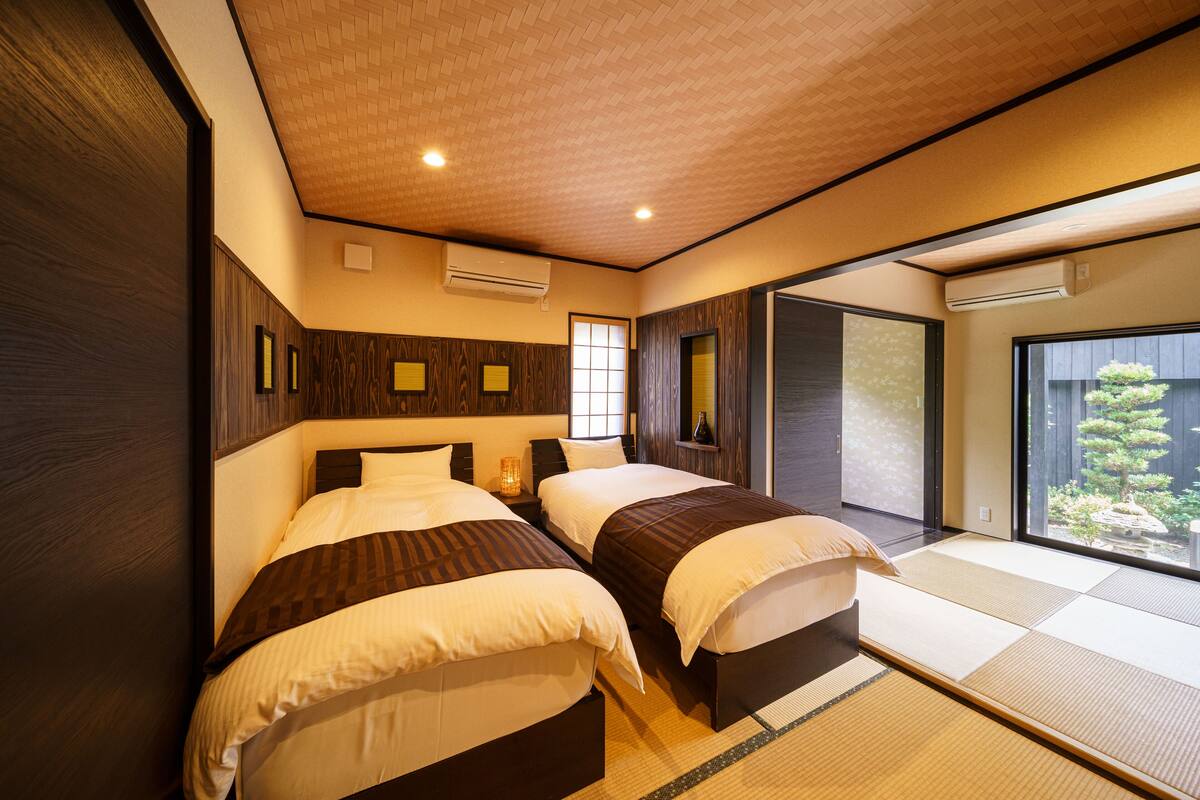 Quarto de hotel com duas camas, painéis de madeira, uma janela grande e vista para um jardim.