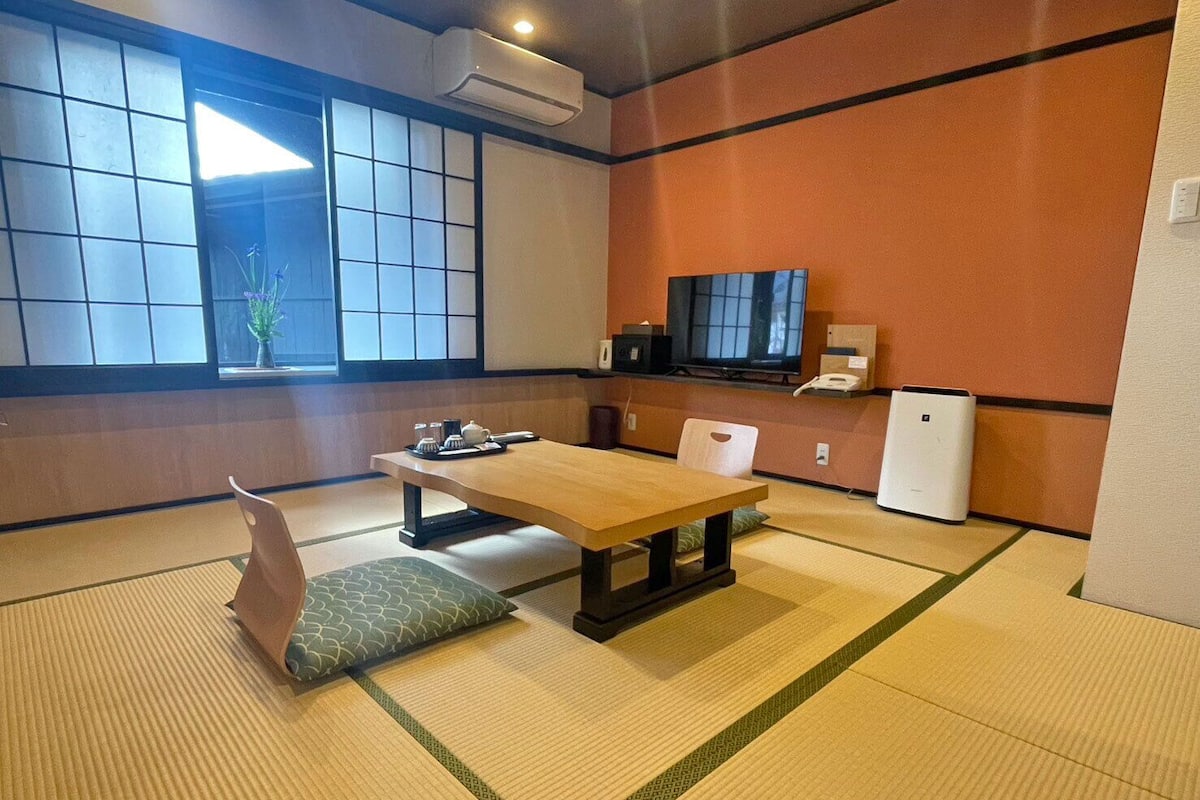 Um quarto tradicional em estilo japonês, com piso de tatame, uma mesa baixa e uma televisão na parede.