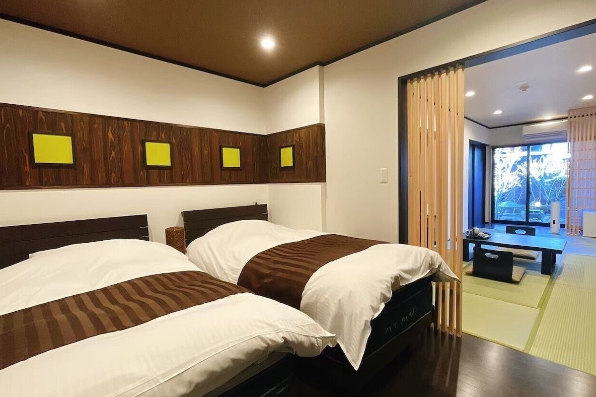Quarto de hotel com duas camas, cabeceira de madeira, uma mesa pequena e vista pela janela.