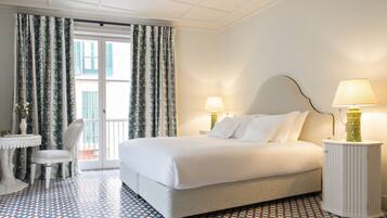 Quarto de hotel com uma cama grande, duas luminárias de cabeceira, uma cadeira, uma mesinha e ladrilhos no chão com padrão.