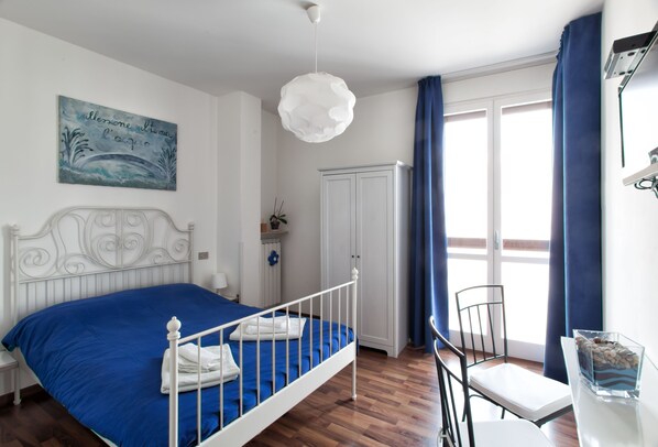 Quarto com estrutura de cama em metal branco, colcha azul, radiador branco, armário branco, cadeira branca, mesa branca e cortina azul.