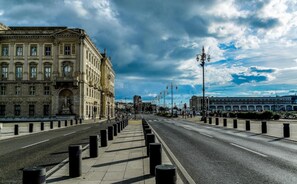 Uma rua larga com calçada, delimitada por defensas, com um grande edifício clássico à esquerda e outro à direita.