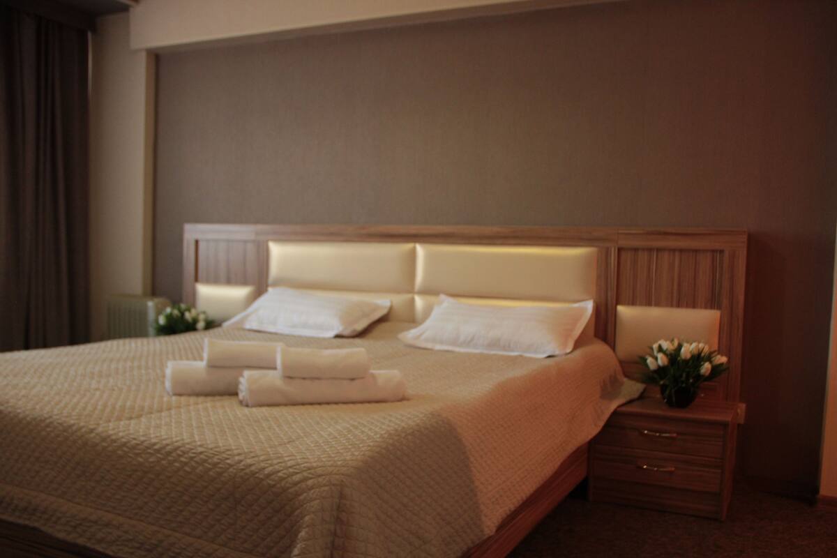 Cama bem arrumada com um travesseiro grande e dois menores, mesa de cabeceira com um vaso de flores e cabeceira de madeira.