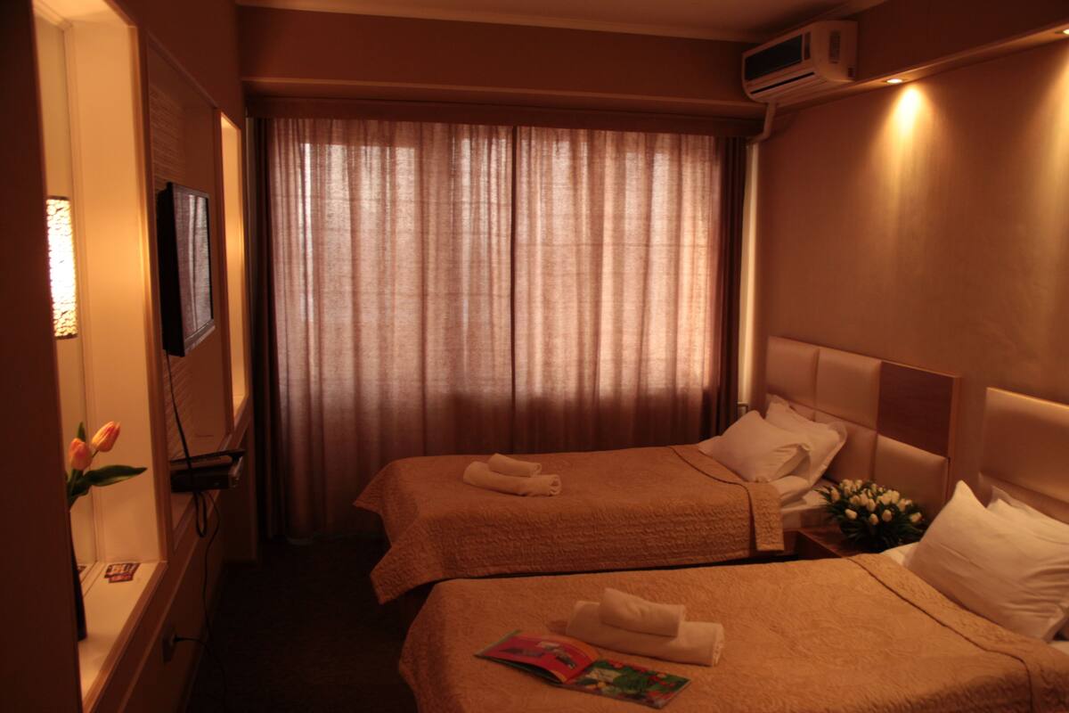 Quarto de hotel com duas camas, televisão, aparelho de ar condicionado e janela com cortinas.