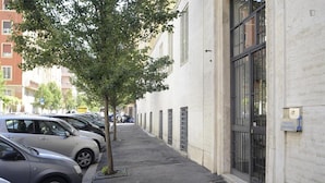 Fila de carros estacionados ao lado de uma calçada, próxima a um edifício com uma grande janela e uma placa.