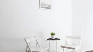 Quarto minimalista com paredes brancas, uma mesinha redonda com planta em vaso e duas cadeiras brancas.