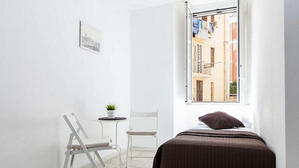 Um quarto pequeno e iluminado, com uma cama, uma cadeira, uma mesinha com uma planta e uma janela com vista para edifícios.