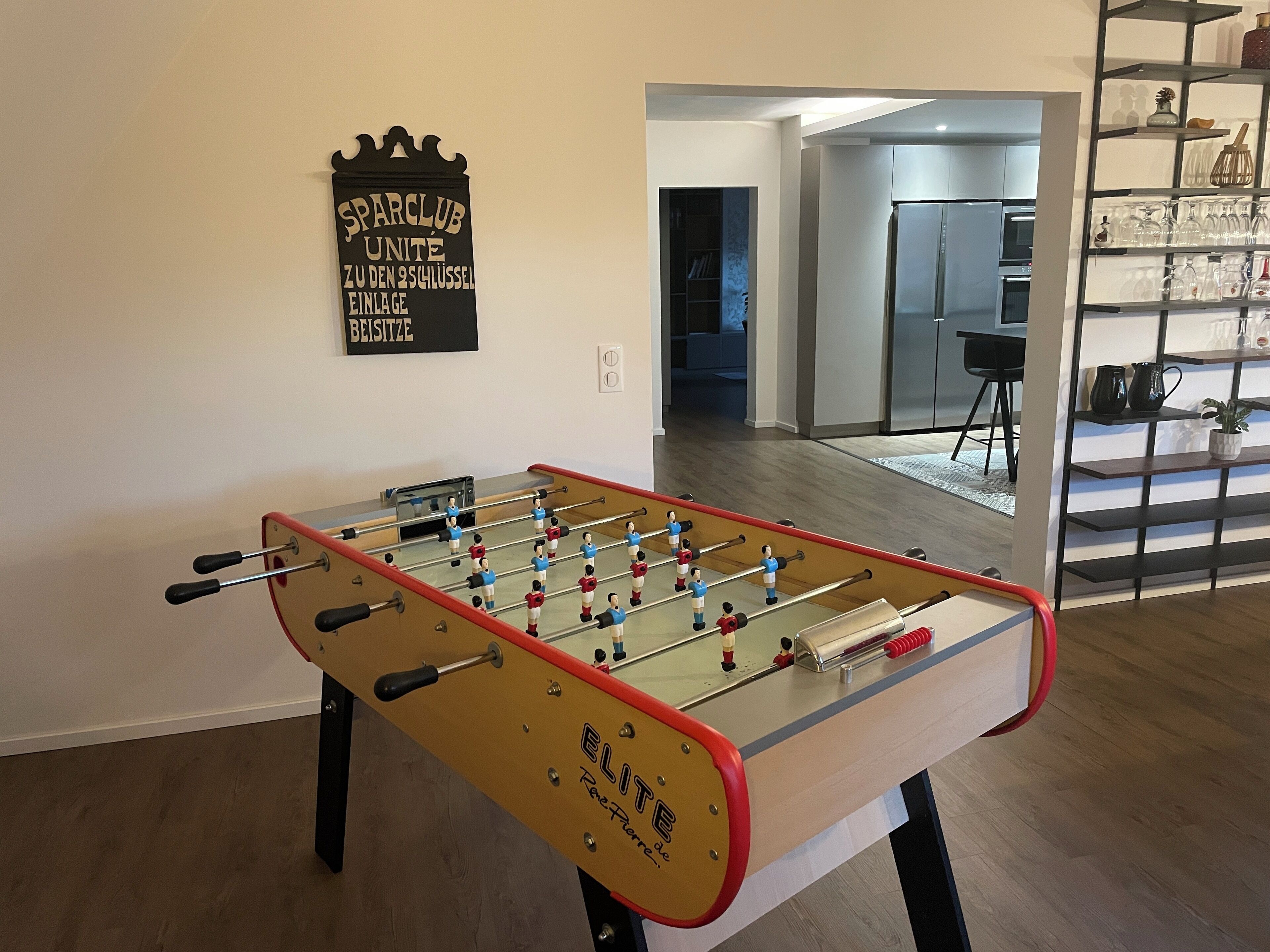 Uma mesa de futebol de mesa em um quarto, com uma área de cozinha visível ao fundo.