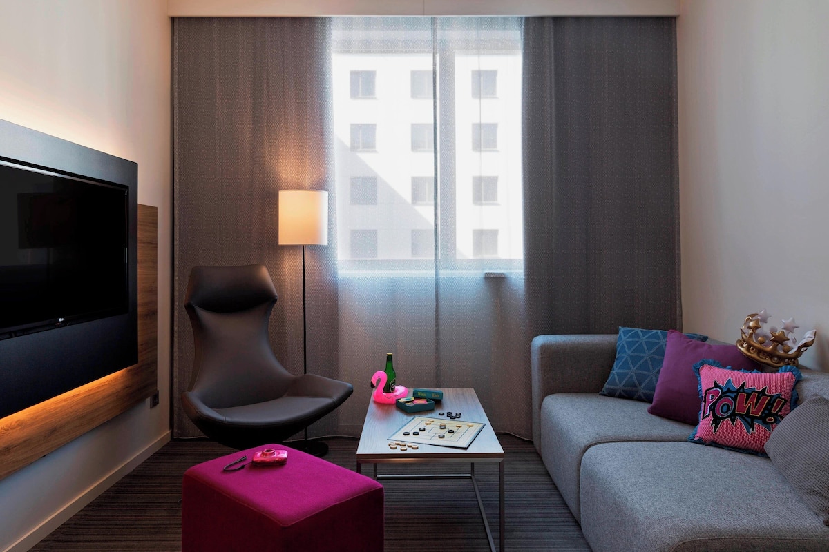 Quarto de hotel moderno com televisão de tela plana, um sofá confortável, uma mesinha de centro com jogos de tabuleiro e uma janela grande com vista para a cidade.