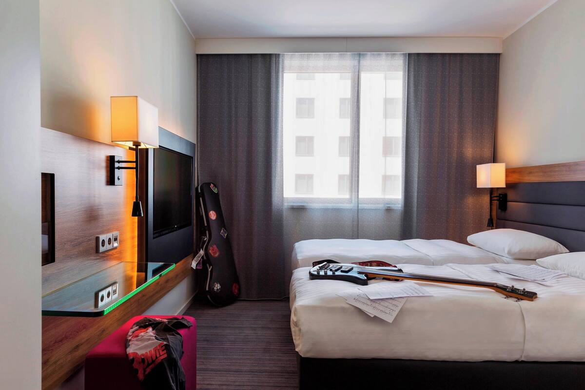 Quarto de hotel com uma cama, uma televisão, um violão e uma mala.