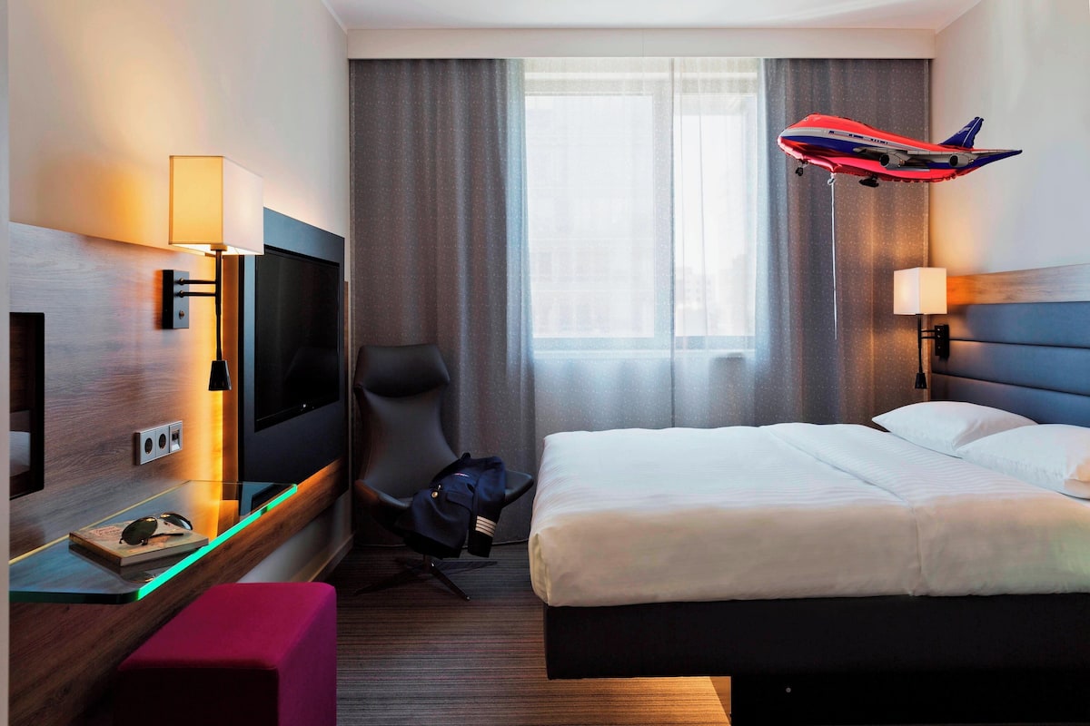 Quarto de hotel com cama, escrivaninha com luminária, cadeira, televisão e janela com cortinas.