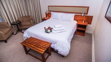 Quarto de hotel com uma cama grande, um banco de madeira, uma cadeira, uma mesinha de cabeceira e um abajur.