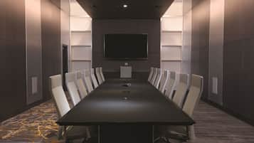 Uma sala de conferências com uma mesa comprida, cadeiras brancas, uma tela grande e piso coberto de carpete.