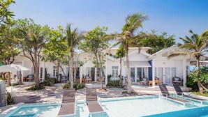 Uma área de piscina com espreguiçadeiras, palmeiras e um edifício branco com varandas.