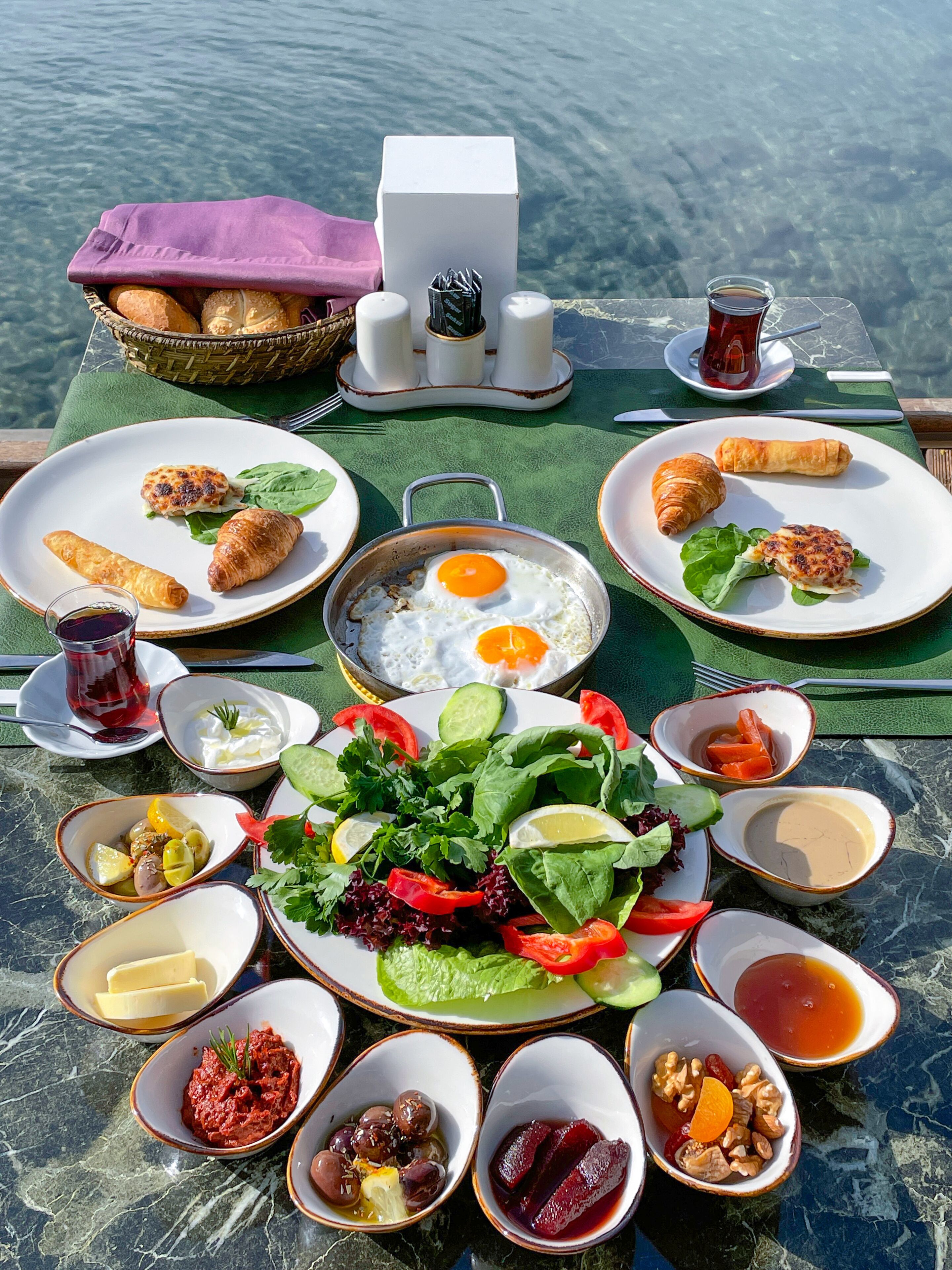 Mesa posta com diversos pratos, incluindo ovos, croissants e saladas, com vista para a água ao fundo.