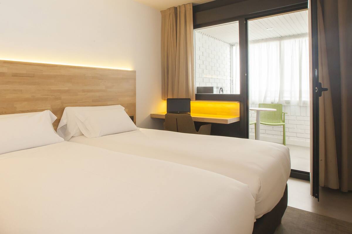 Quarto de hotel com uma cama grande, uma escrivaninha com um laptop e vista para a área externa.