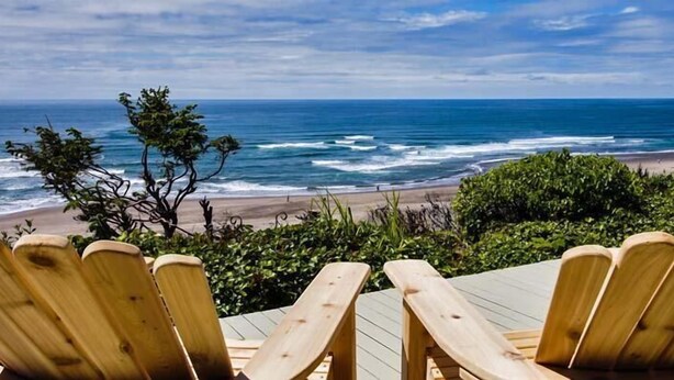 Um deck de madeira com duas cadeiras Adirondack voltadas para uma praia com ondas e céu claro.