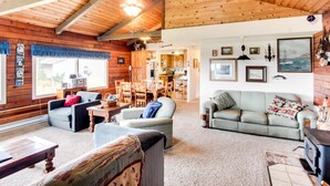 Interior acolhedor de uma cabana, com paredes de madeira, área de jantar e sala de estar com sofá e poltronas.