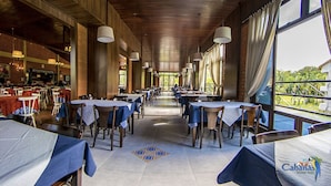 Uma sala de jantar ampla, com mesas e cadeiras, janelas grandes e vista para o verde lá fora.
