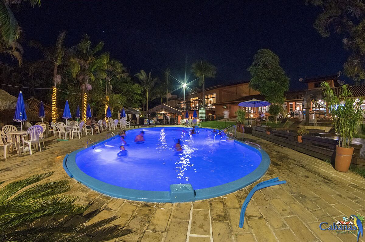 Uma área de piscina à noite, com espreguiçadeiras e guarda-sóis, cercada por palmeiras e um edifício iluminado.