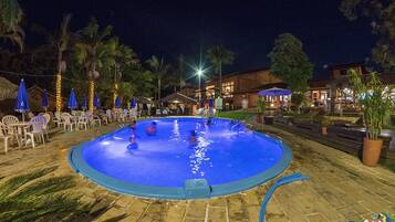 Uma área de piscina à noite, com espreguiçadeiras e guarda-sóis, cercada por palmeiras e um edifício iluminado.
