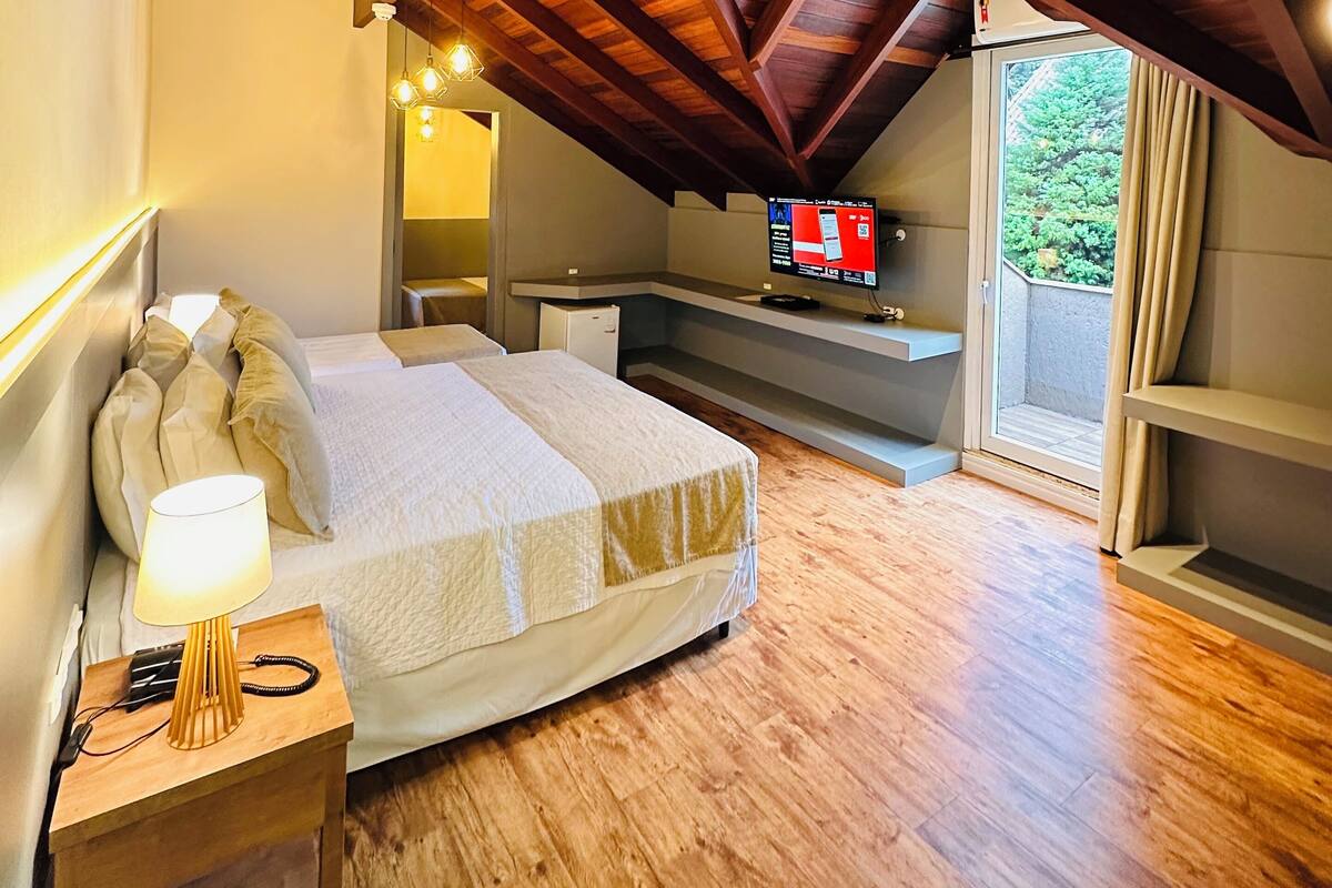 Quarto com teto de madeira, uma cama, mesa de cabeceira com abajur, televisão e janela com cortinas.