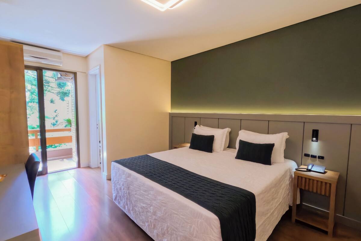 Quarto moderno com uma cama grande, mesa de cabeceira com abajur, e vista para o exterior através de uma porta deslizante.