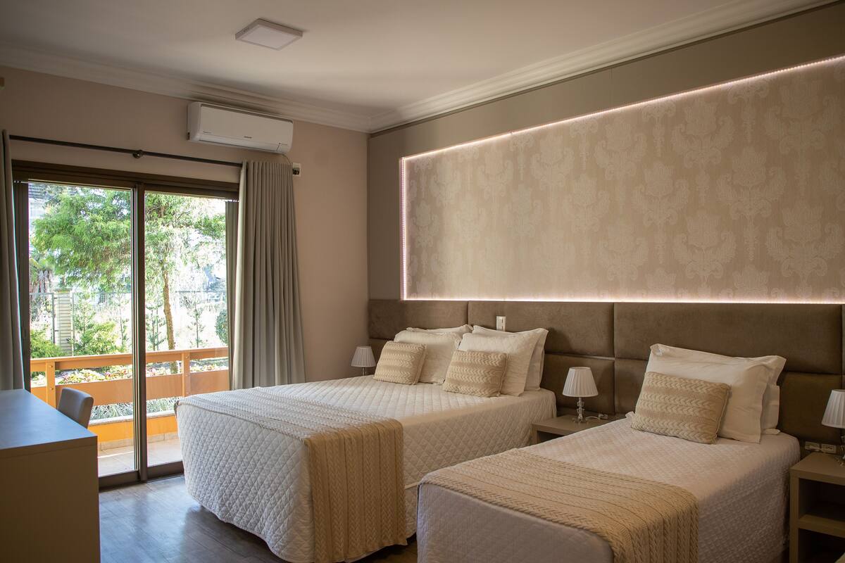 Quarto de hotel com duas camas, uma escrivaninha e varanda com vista para árvores.