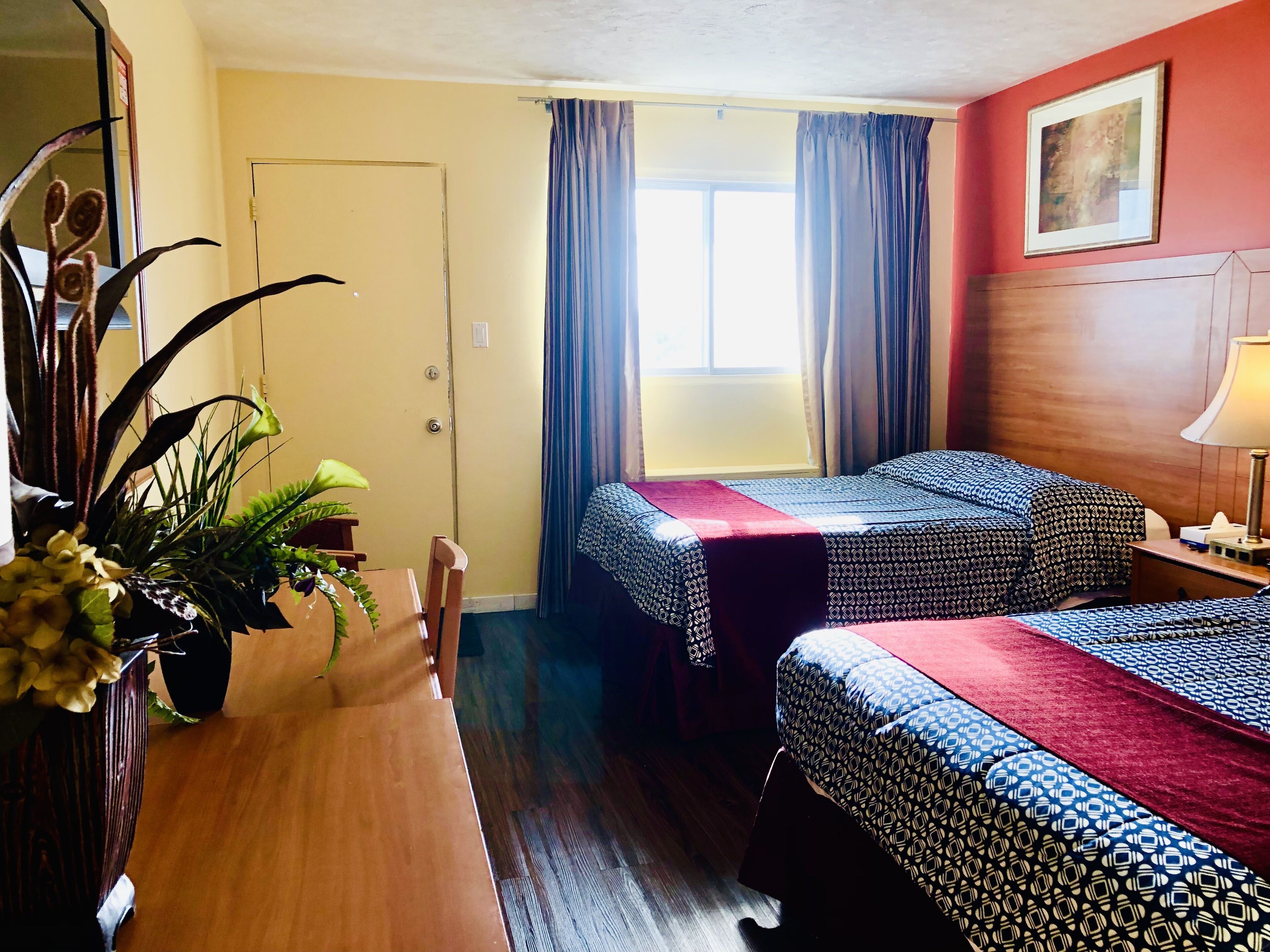 Quarto de hotel com duas camas, uma escrivaninha de madeira, um vaso de plantas e uma janela com cortinas.
