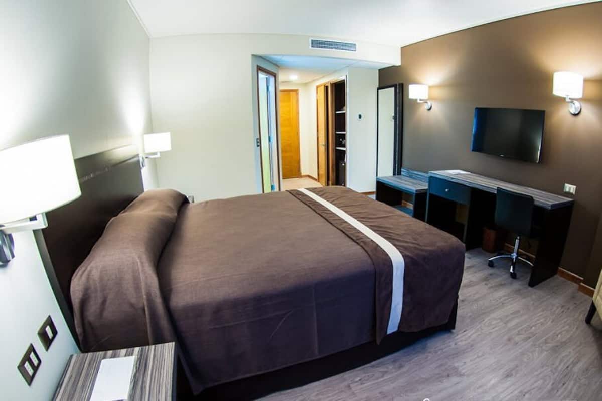Quarto de hotel com uma cama grande, uma escrivaninha com cadeira, televisão e guarda-roupa.